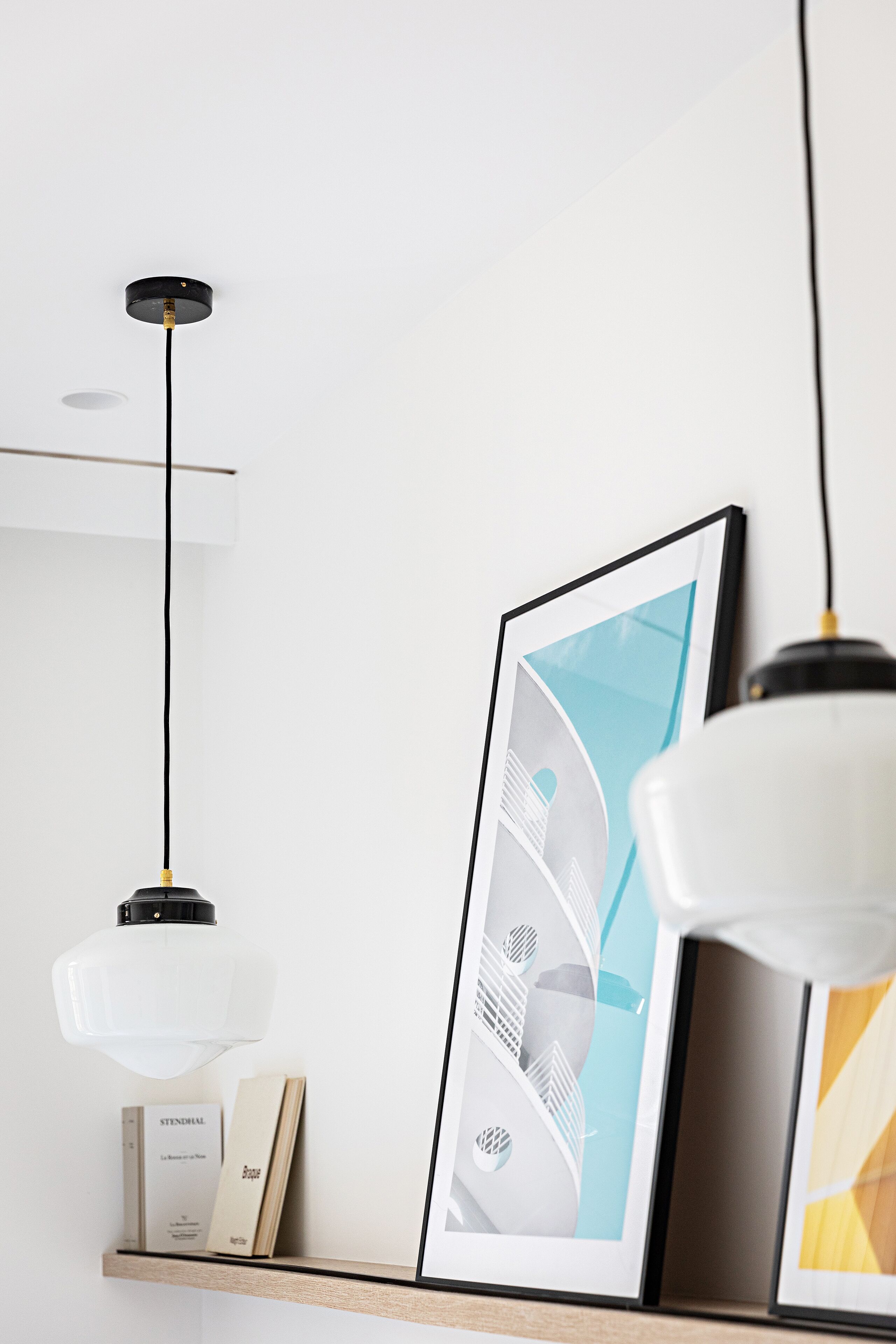 Um interior moderno com teto branco, prateleira de madeira com livros e uma obra de arte abstrata emoldurada, e duas luminárias pendentes.