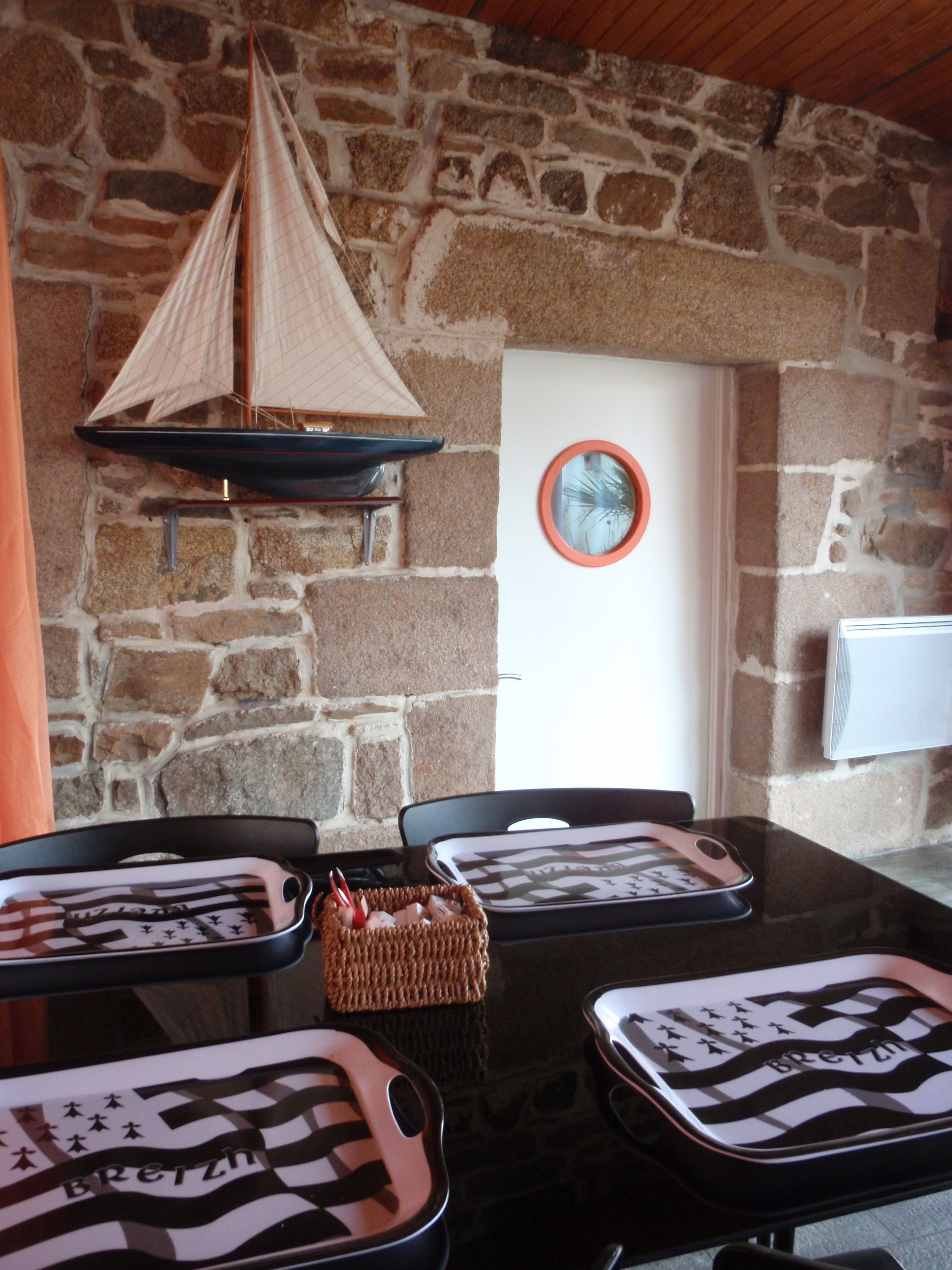 Quarto com paredes de pedra e teto de madeira, um modelo de veleiro pendurado, um espelho redondo e uma mesa com bandejas.