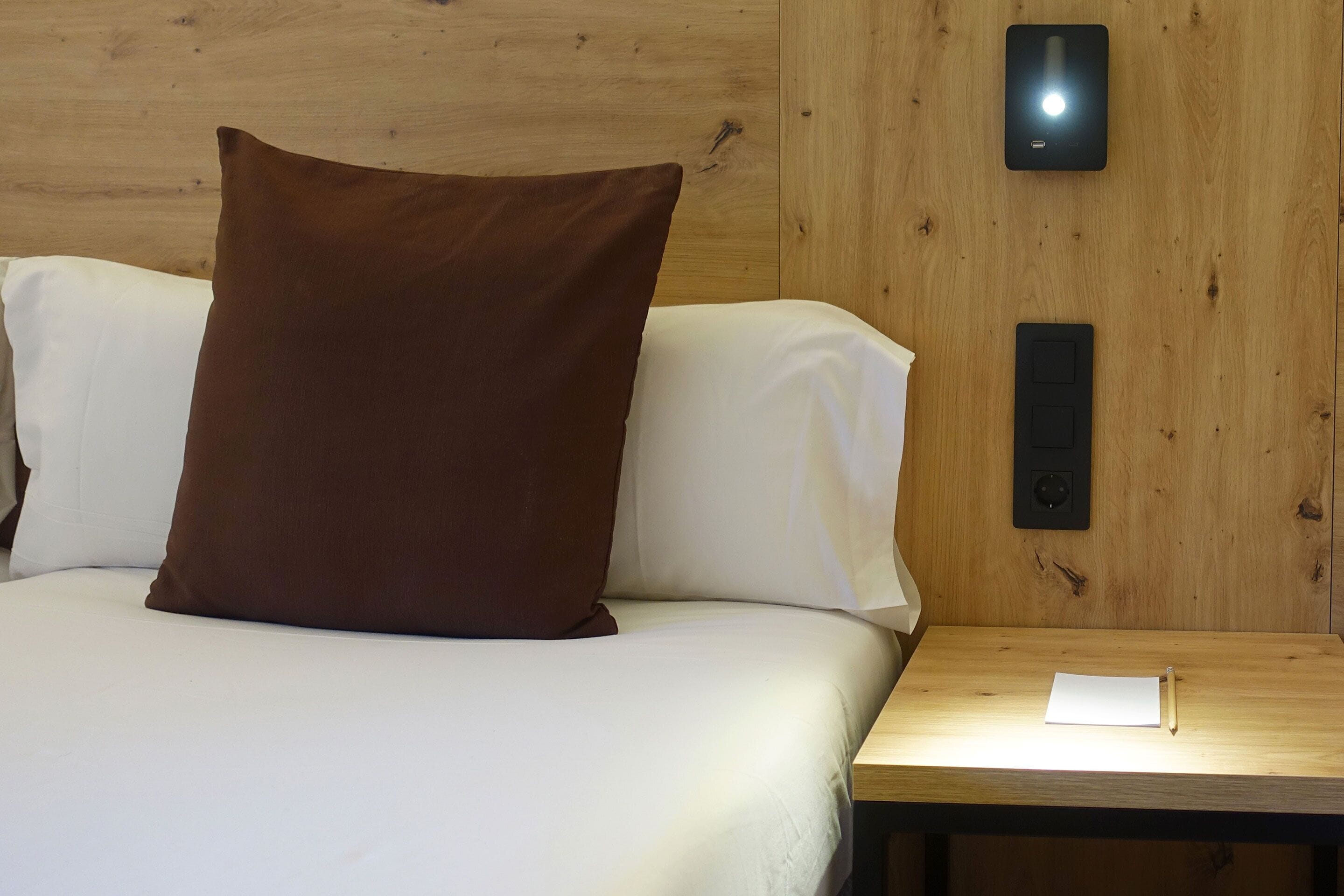 Cama bem arrumada com roupa de cama branca e castanha, mesa de cabeceira com abajur e painel de controle fixado na parede.