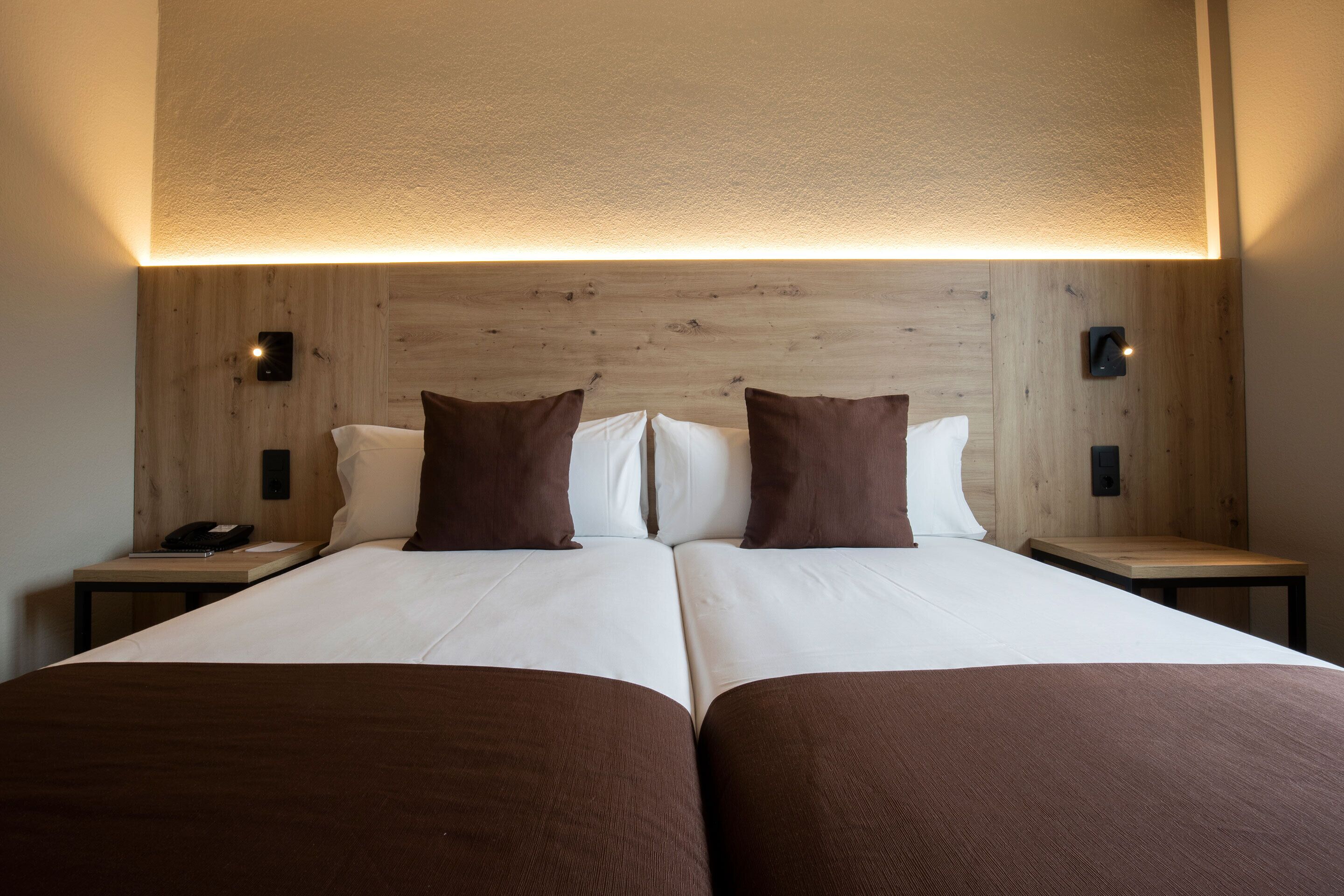 Uma cama de casal com duas almofadas, mesa de cabeceira com telefone e cabeceira de madeira com luzes na parede.