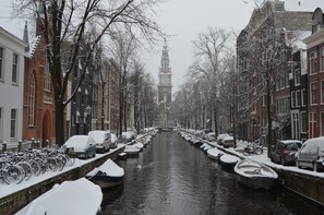 Um canal flanqueado por carros e bicicletas cobertos de neve, com edifícios em ambos os lados e uma torre ao fundo.