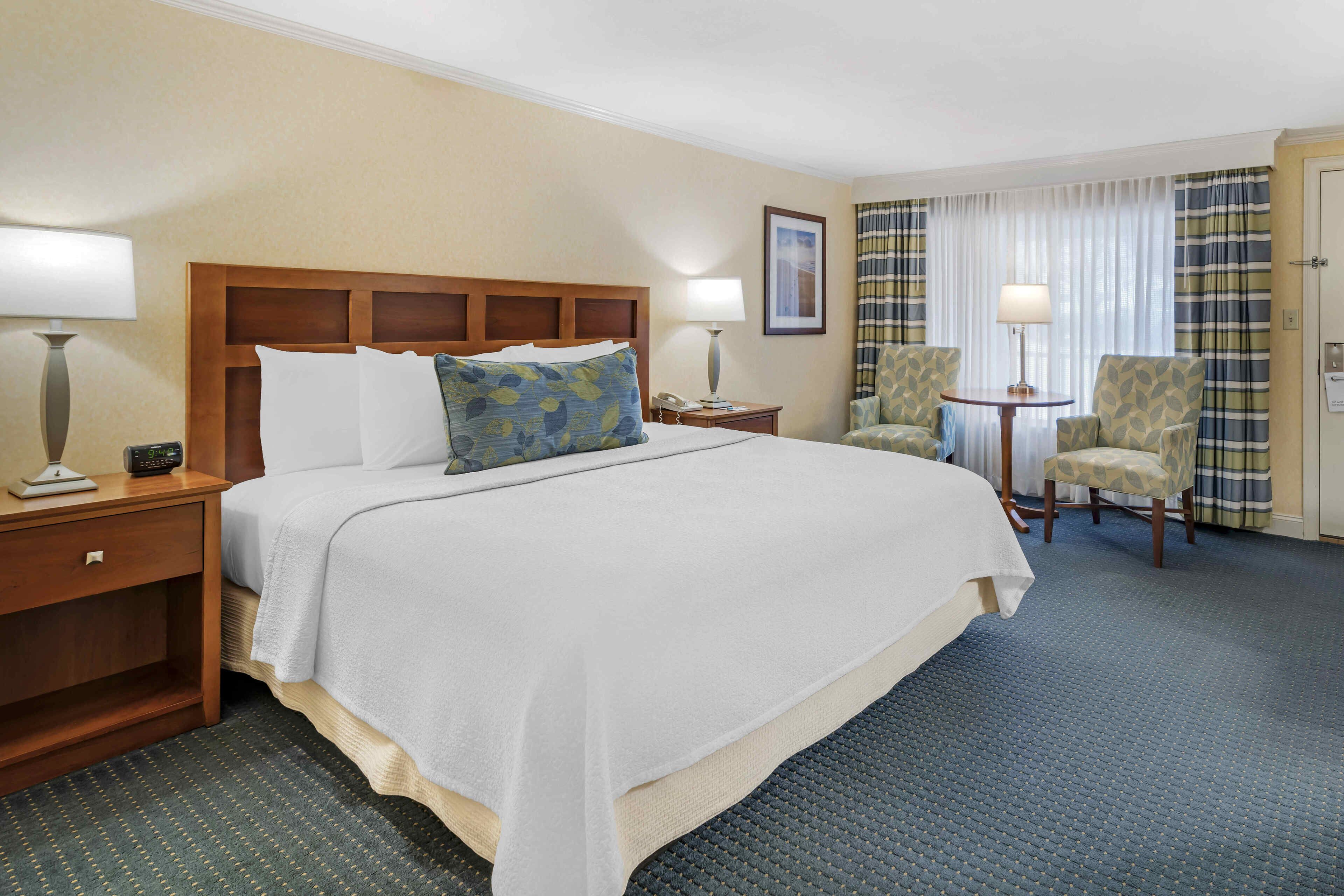 Quarto de hotel com uma cama grande, duas mesas de cabeceira com abajures, uma cadeira e uma pequena mesa com luminária.