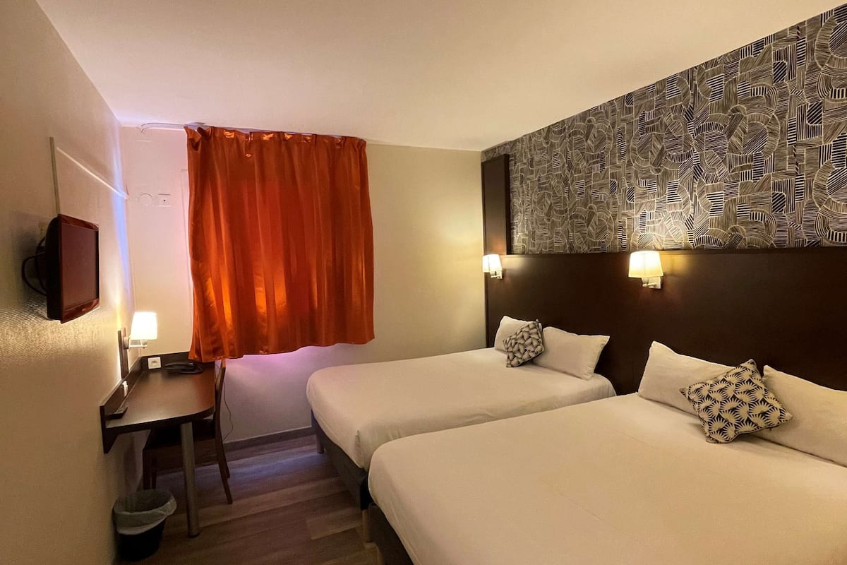 Quarto de hotel com duas camas, uma escrivaninha, uma cadeira, um cesto de lixo e parede com padrão.