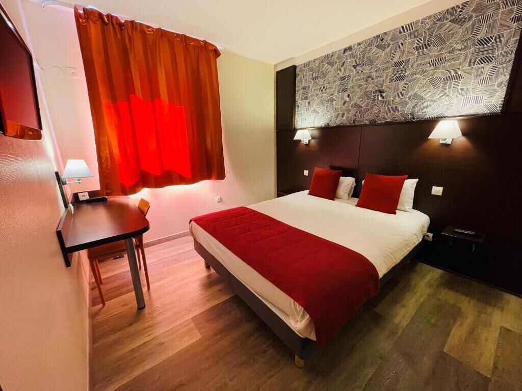 Quarto de hotel com cama, escrivaninha e cadeira. Há uma janela com cortinas, uma parede com padrão geométrico e duas luminárias fixadas na parede.