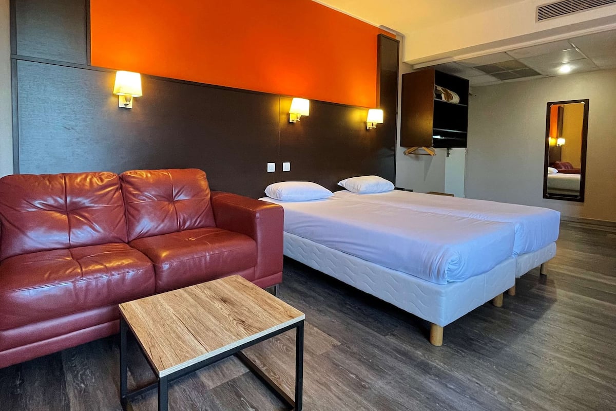 Quarto de hotel com sofá de couro vermelho, duas camas de solteiro, mesa de centro de madeira e luminárias fixadas na parede.