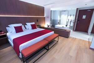 Quarto de hotel moderno com uma cama grande, um banco, uma escrivaninha, toucador com espelho e banheiro com banheira e toalhas.