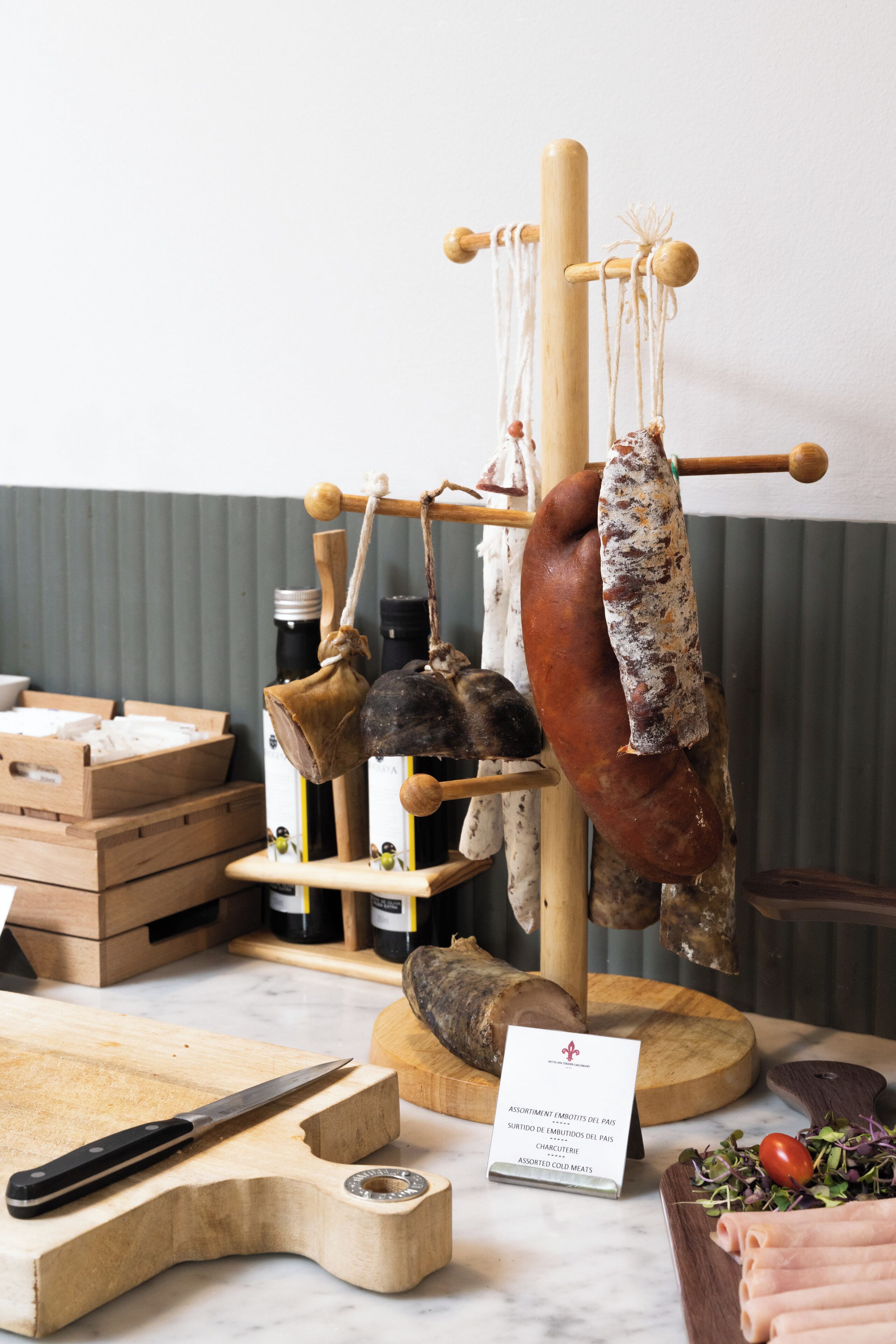 Exposição de diversos tipos de carne, incluindo um linguiça pendurada, em um suporte de madeira com uma placa indicando 'Assortimento de Frios'.