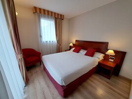 Quarto de hotel com uma cama grande, duas mesas de cabeceira com luminárias, uma cadeira vermelha e uma janela com cortinas.
