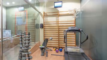 Academia com esteira, aparelhos de musculação, um banco e uma TV fixada na parede.