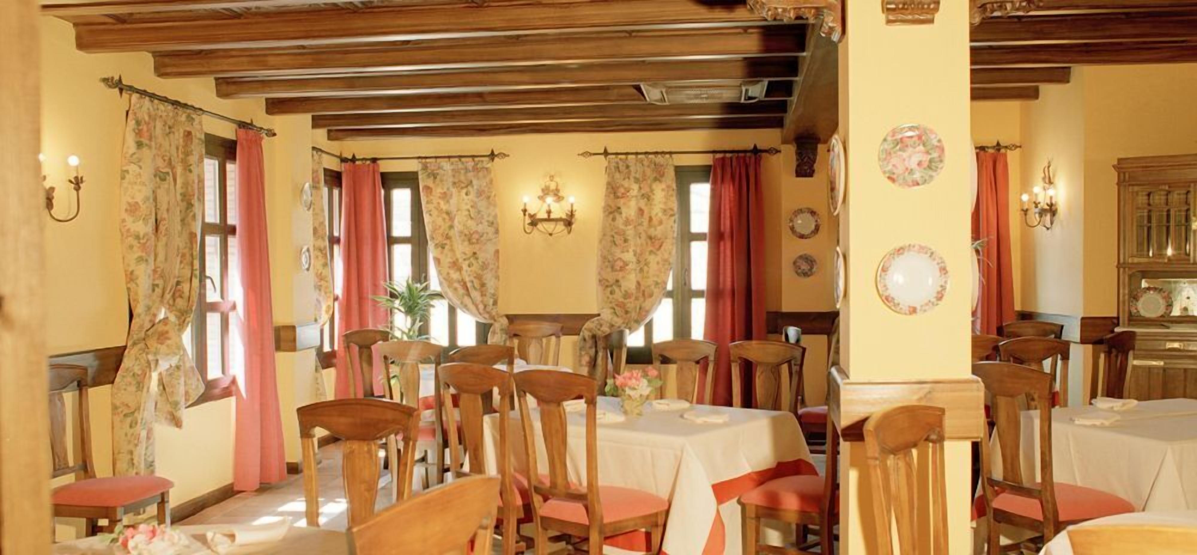 Uma sala de jantar com mesas e cadeiras de madeira, cortinas florais e luminárias fixadas na parede.