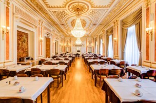 Uma imponente sala de banquetes com fileiras de mesas e cadeiras, detalhes ornamentais no teto e um grande candelabro.