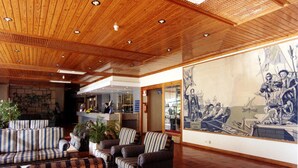 Um hall com teto de madeira, sofás estampados, balcão de recepção e um grande mural histórico.
