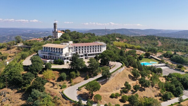 Um edifício grande, com vários andares e uma torre imponente, cercado por vegetação exuberante e uma estrada sinuosa.