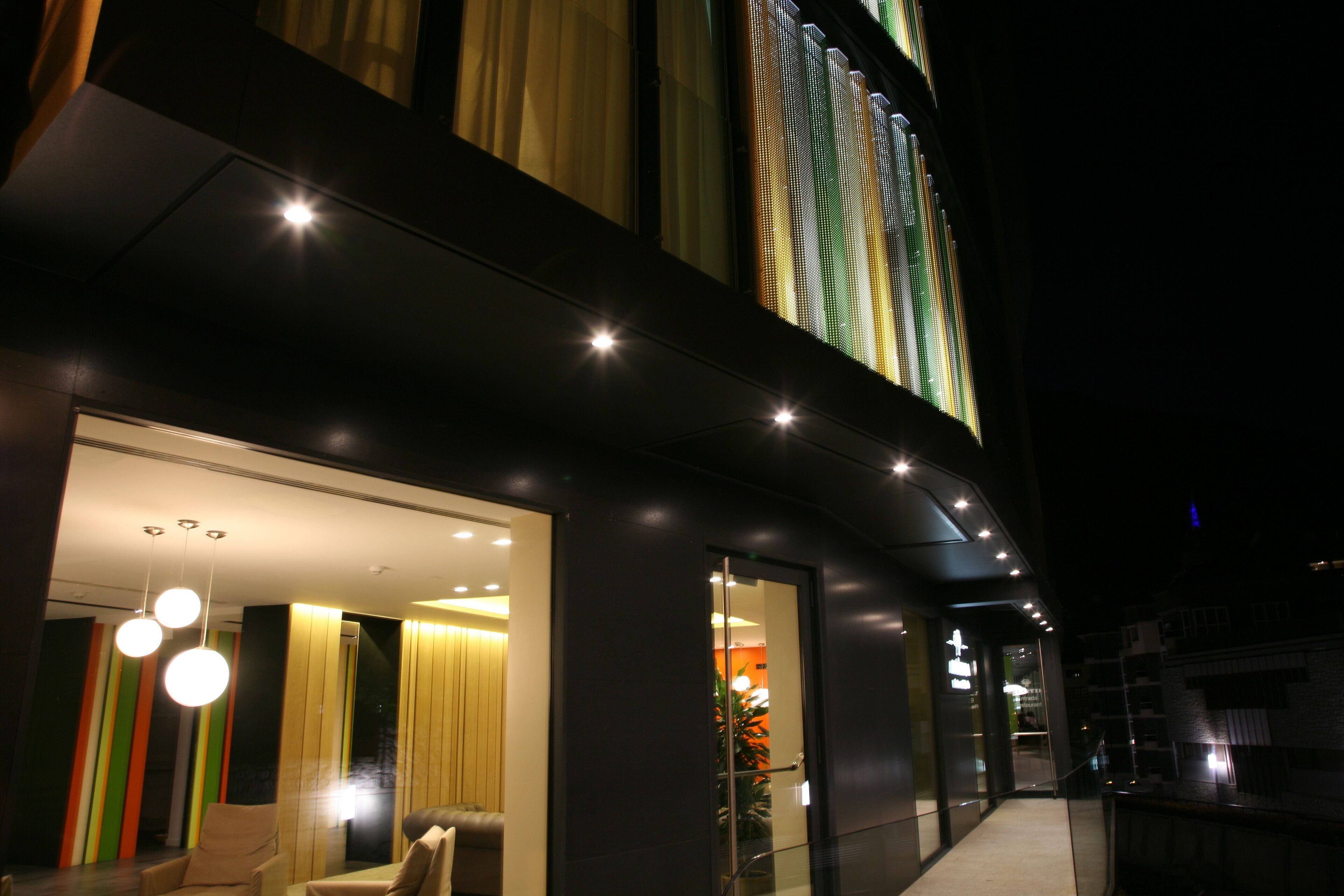 Exterior moderno de hotel à noite, com janelas iluminadas, entrada de vidro e escadaria visível.
