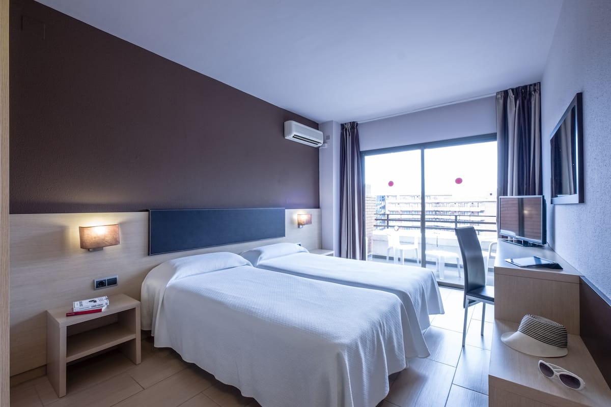 Quarto de hotel com duas camas, uma escrivaninha com cadeira, televisão e varanda com vista.