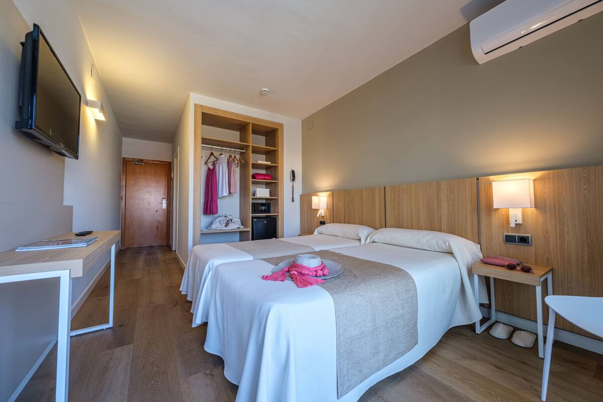 Quarto de hotel com duas camas, televisão, escrivaninha, guarda-roupa e mesa de cabeceira com chinelos.