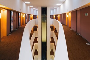 Corredor de hotel com elevador central, várias portas de ambos os lados e piso coberto por carpete.