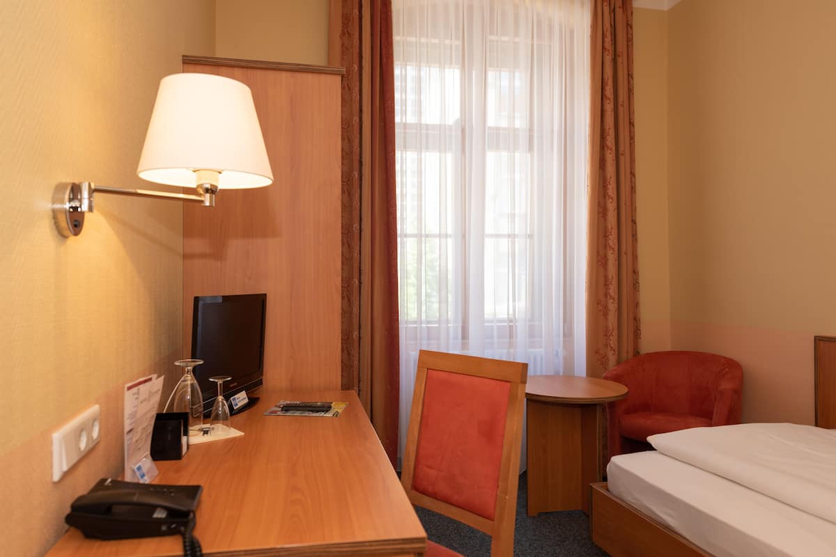 Quarto de hotel com escrivaninha de madeira, televisão, cama, cadeira e janela com cortinas.