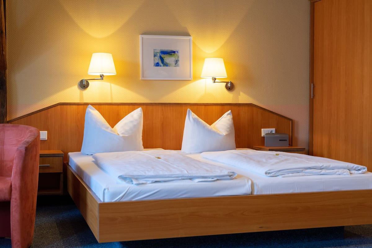 Cama de casal com roupa de cama branca, duas luminárias de cabeceira, uma obra de arte emoldurada acima da cama e cabeceira de madeira.