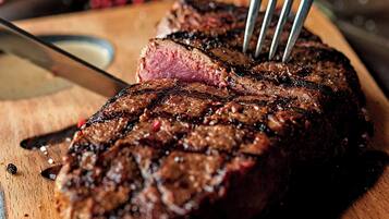 Bife sendo grelhado em uma superfície de madeira, com faca e garfo.