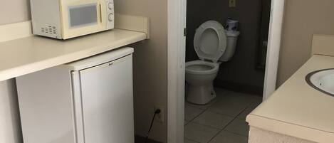 Uma kitchenette compacta com micro-ondas, máquina de café e pequena geladeira. Um vaso sanitário é visível no banheiro adjacente.