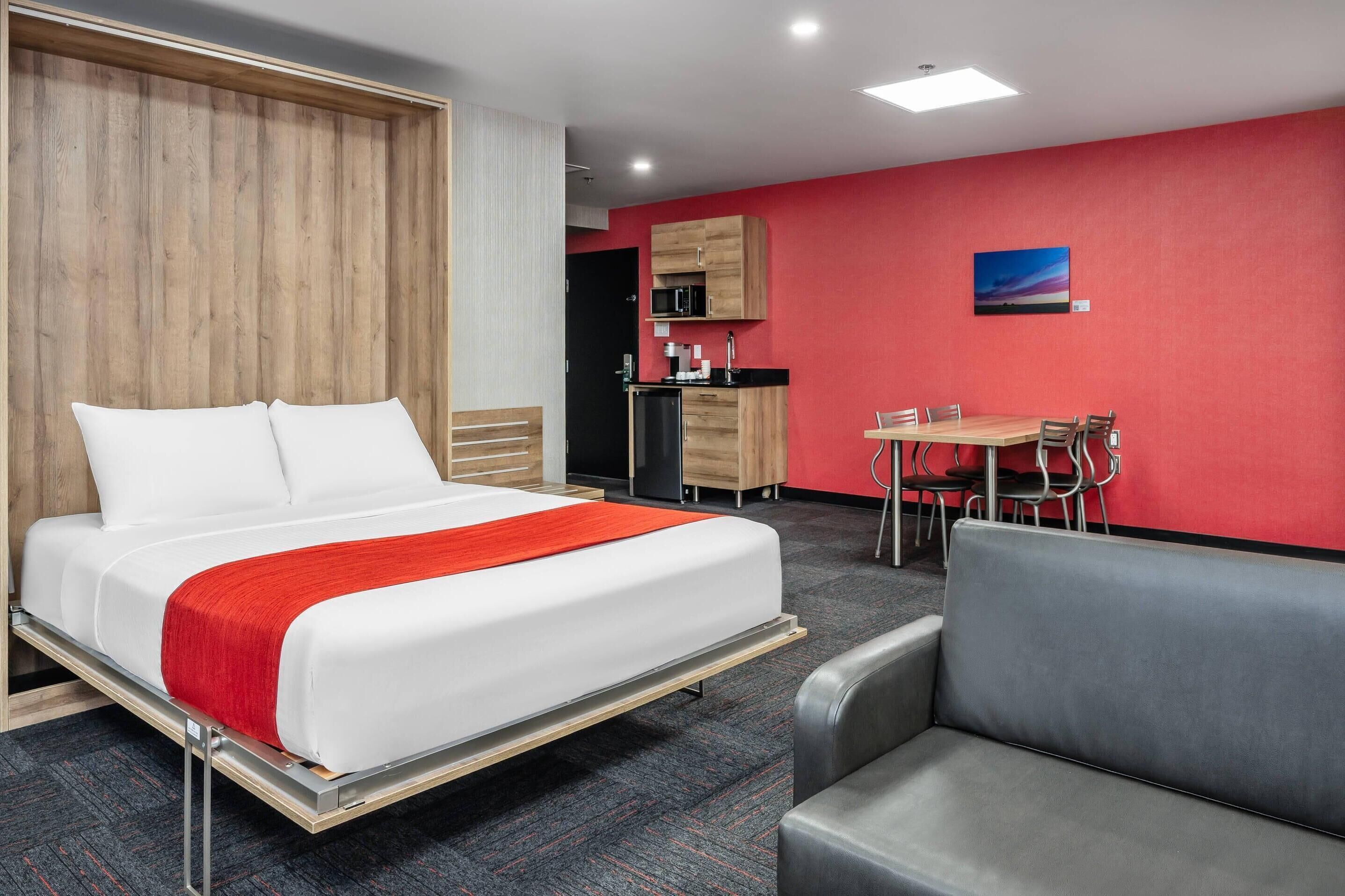 Quarto de hotel com cama, sofá cinza, mesa de jantar com cadeiras, cozinha compacta e parede vermelha.