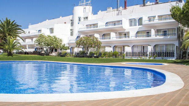 Piscina com deck de madeira, rodeada por um edifício branco com varandas e um céu azul límpido.