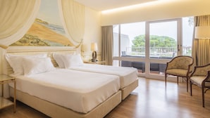 Quarto de hotel com duas camas, uma janela grande com vista para a praia e uma cadeira.