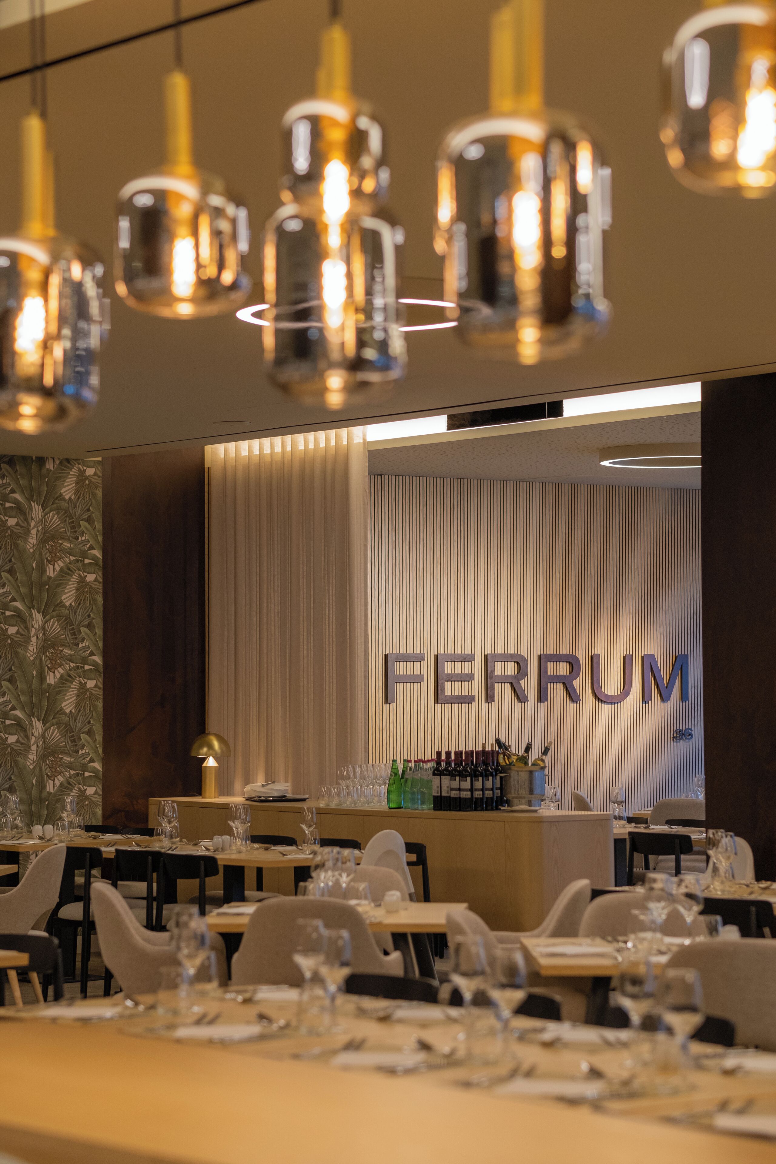 Interior de restaurante com mesas preparadas para refeições, luminárias pendentes e uma parede com a palavra 'FERRUM'.
