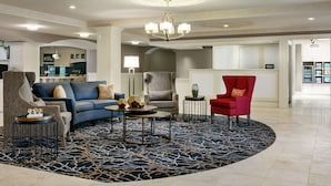 Um hall amplo com carpete padronado azul, um candelabro e sofás, incluindo uma poltrona vermelha e uma poltrona cinza.