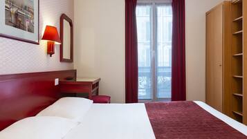 Quarto de hotel com cama, escrivaninha, cadeira, abajur, guarda-roupa e janela com cortinas.