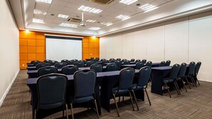 Uma sala de conferência com mesas e cadeiras dispostas em fileiras, uma tela de projetor e um quadro branco.