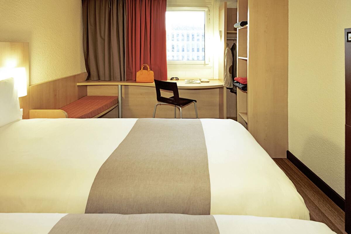 Quarto de hotel com duas camas, uma escrivaninha, uma cadeira, uma janela com cortinas e um guarda-roupa.