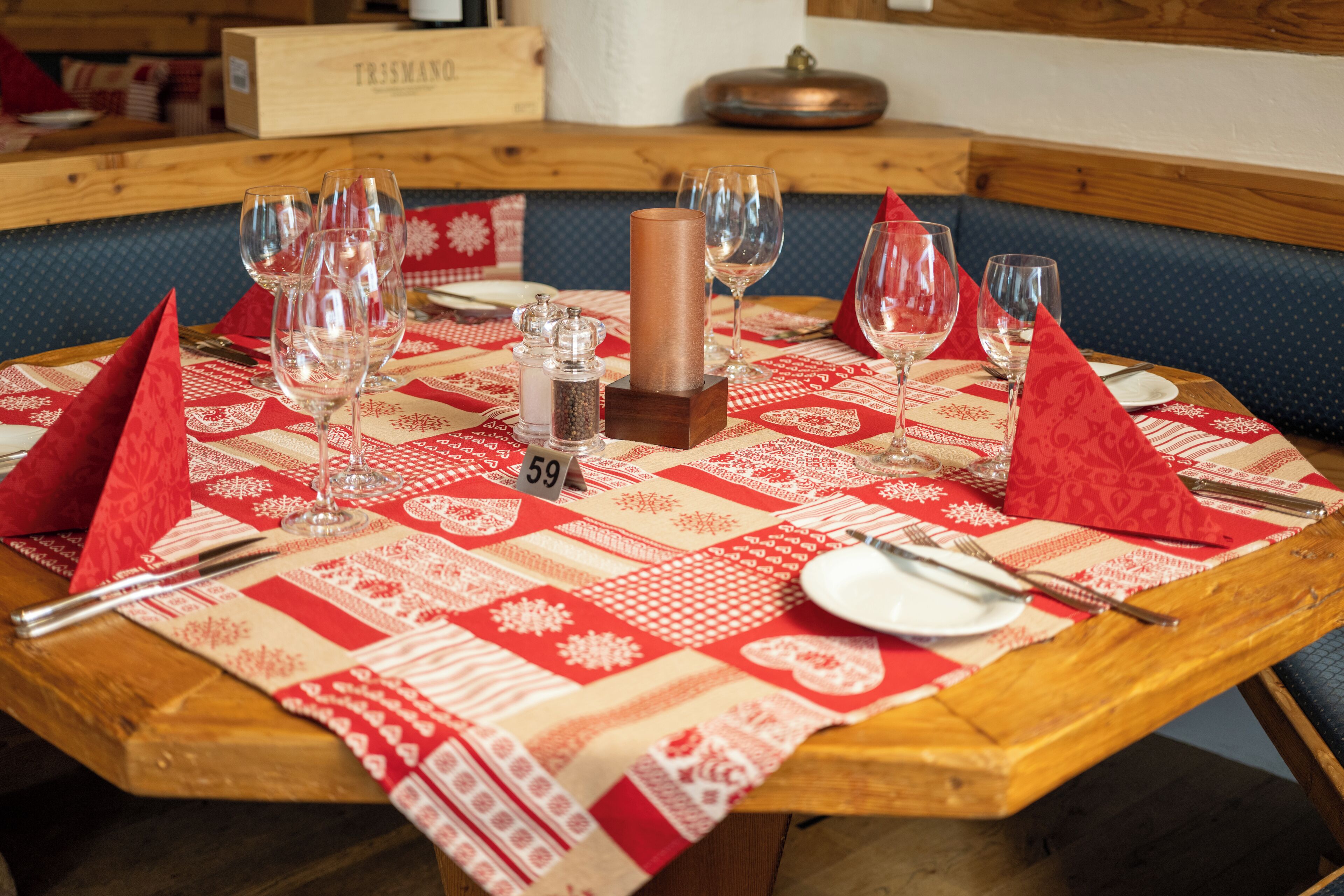 Mesa de jantar posta com copos de vinho, pratos e talheres, coberta por uma toalha de mesa estampada em vermelho e branco.