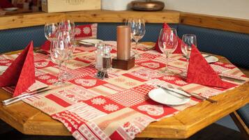 Mesa de jantar posta com copos de vinho, pratos e talheres, coberta por uma toalha de mesa estampada em vermelho e branco.
