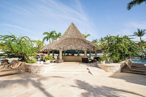 Um pavilhão com telhado de palha, piscina e espreguiçadeiras em um cenário tropical.