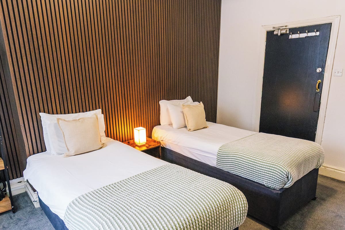 Quarto de hotel com duas camas, parede de ripas de madeira, porta preta e pequena mesa de cabeceira com abajur.