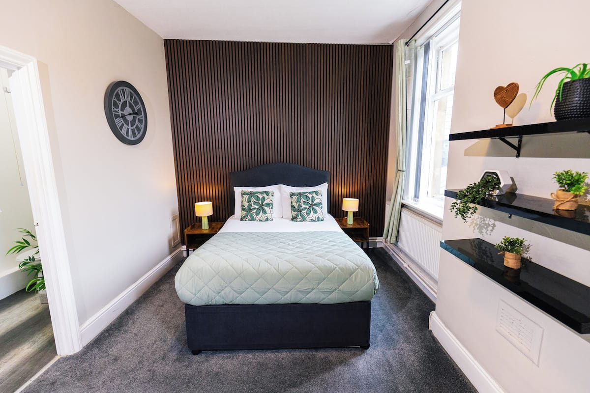 Quarto com cama, luminárias de cabeceira, relógio, janela e prateleira com plantas.