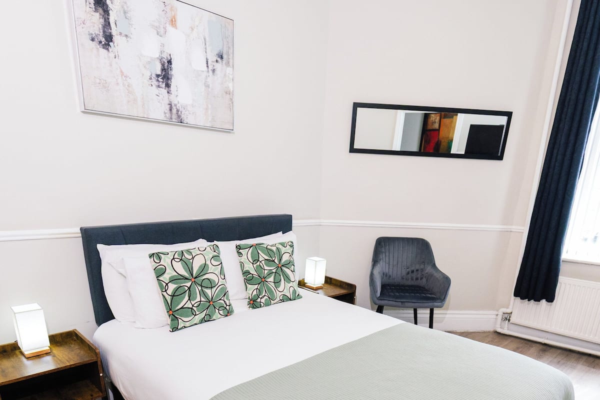 Quarto de hotel bem arrumado, com uma cama, mesas de cabeceira, uma cadeira, um espelho fixado na parede e um quadro.