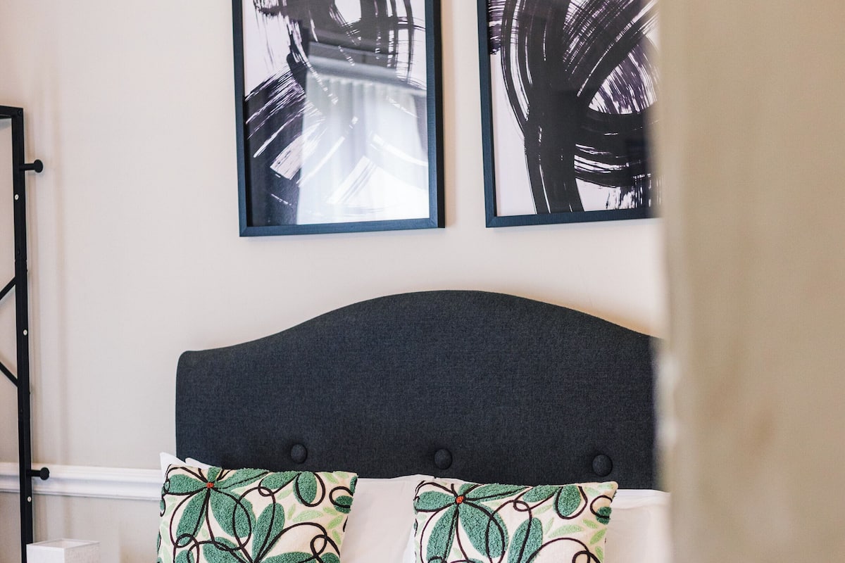 Uma cama com cabeceira escura, duas almofadas estampadas e duas pinturas abstratas na parede.