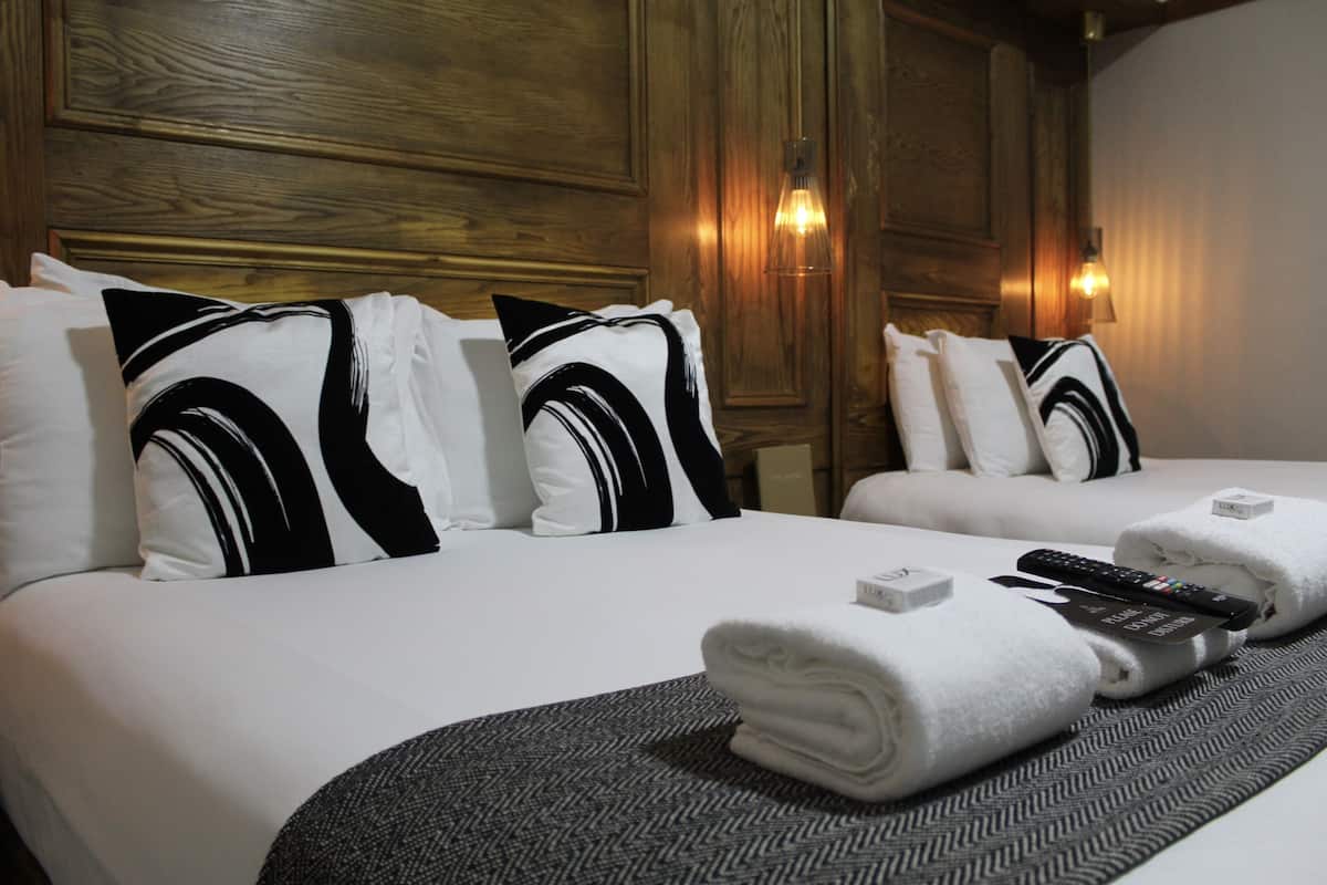 Quarto de hotel com duas camas, cada uma com roupa de cama branca e almofadas preto e branco. Há dois controles remotos e uma toalha dobrada na cama.