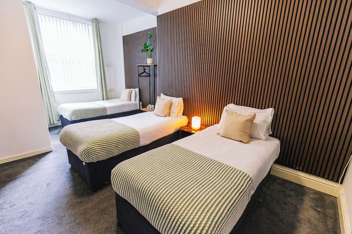 Quarto de hotel com duas camas, uma mesa de cabeceira com luminária, um vaso de planta e uma janela com cortinas.