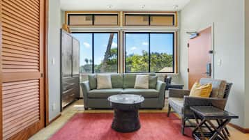 Uma sala de estar com sofá, mesa de centro, cadeira de vime e uma janela grande com vista para árvores e prédios.