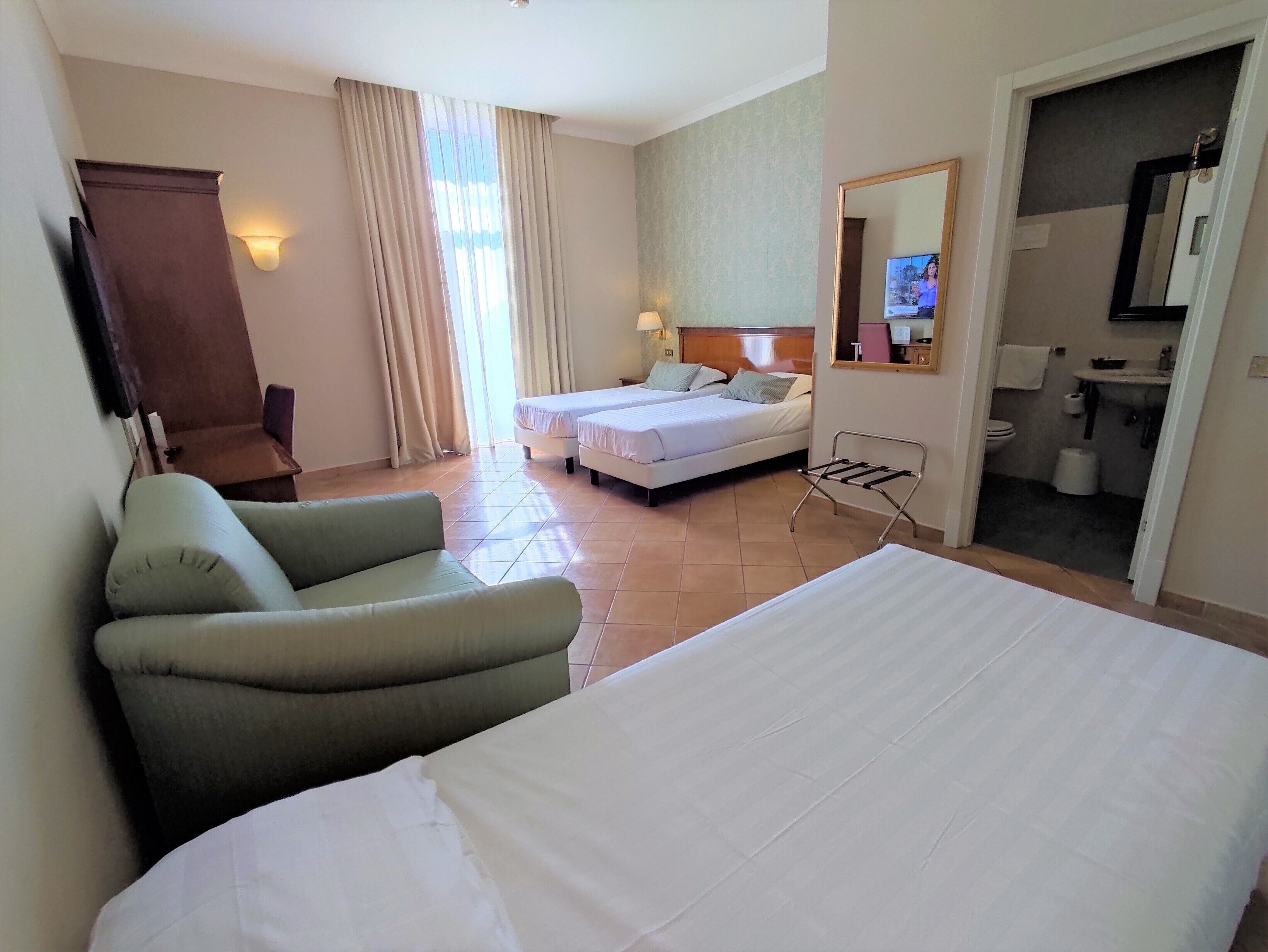 Quarto de hotel com cama, duas mesas de cabeceira, uma cadeira, televisão, banheiro com pia e vaso sanitário, e janela com cortinas.