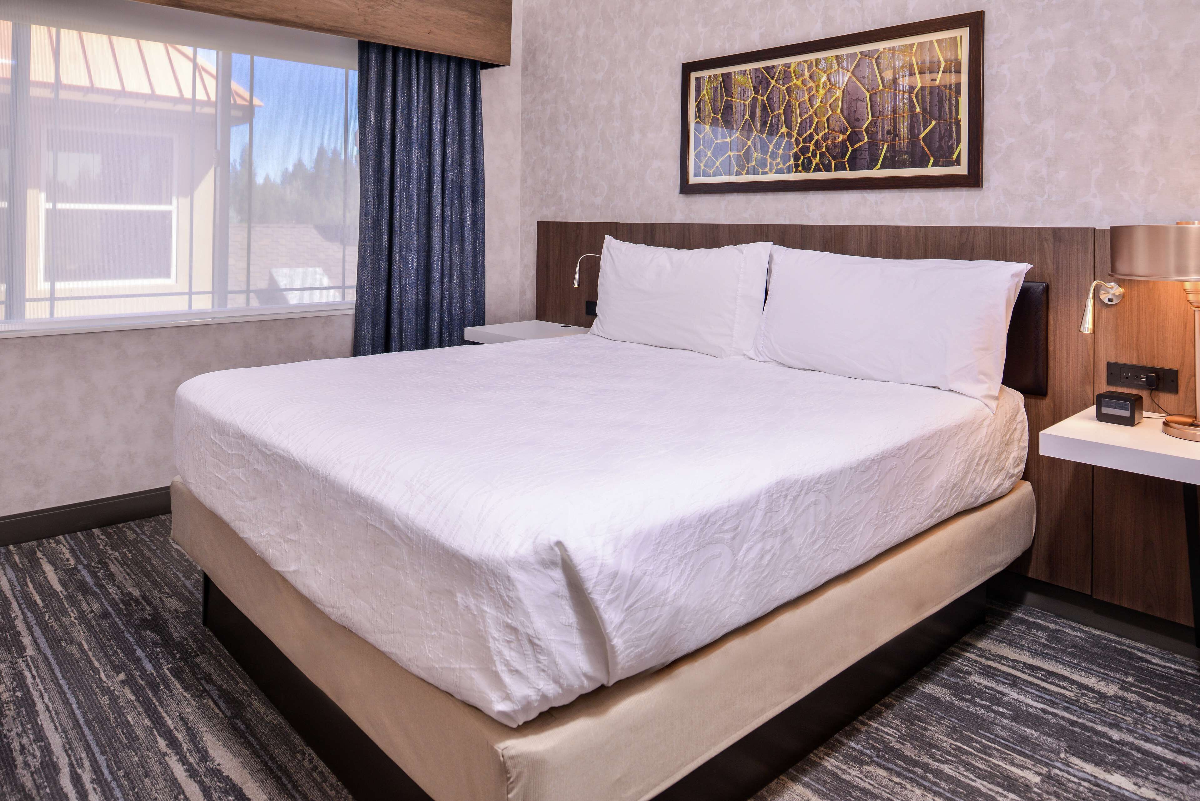 Quarto de hotel com uma cama grande, mesa de cabeceira, abajur e um quadro na parede.