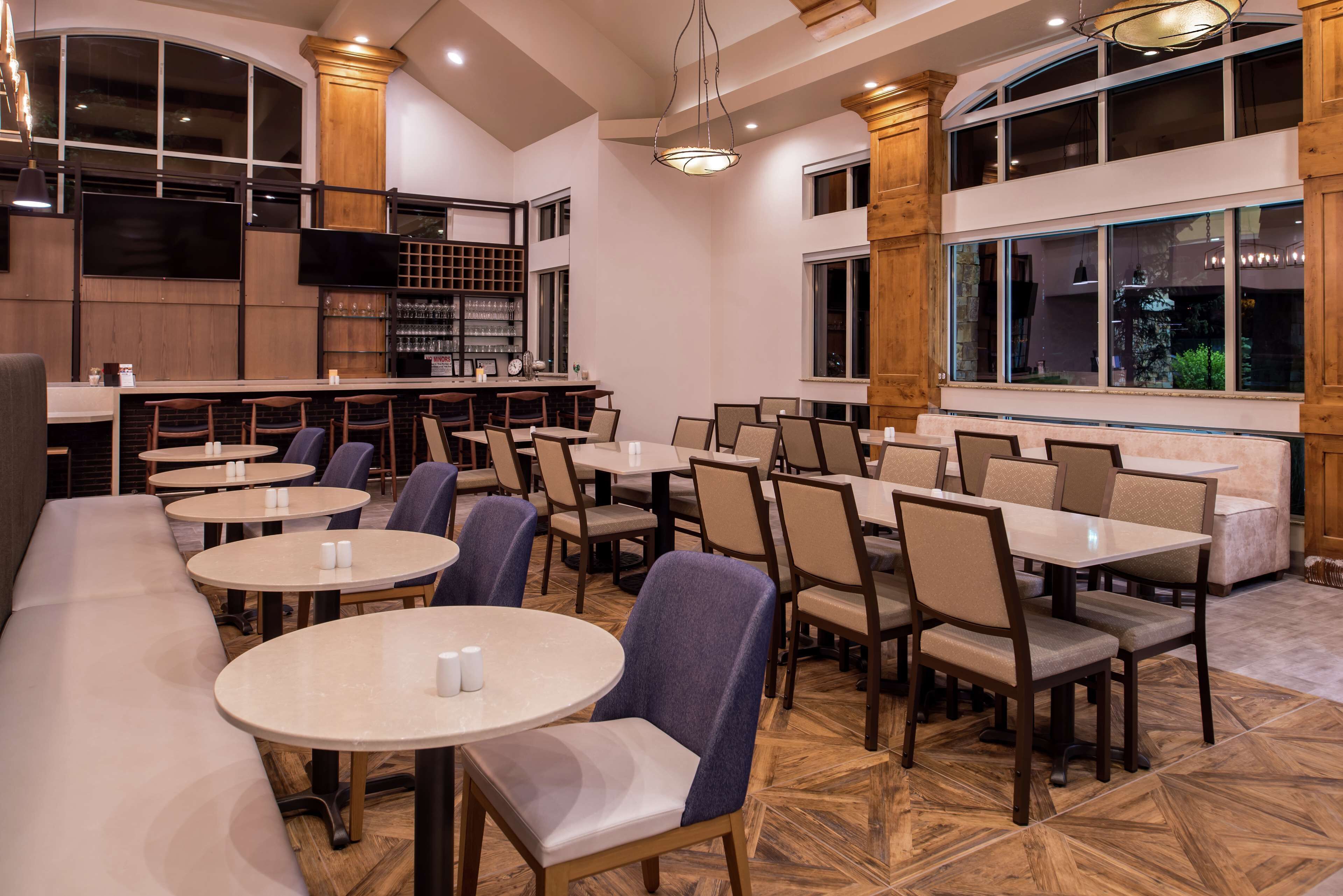 Restaurante com várias mesas e cadeiras redondas, um espaço de bar e amplas janelas que proporcionam luz natural.