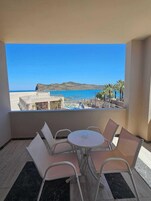 Varanda com mesa e cadeiras, vista para uma praia e céu azul límpido.