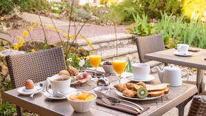 Café da manhã ao ar livre com cadeiras de vime, uma mesa com itens de café e vista para o jardim.