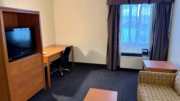 Quarto de hotel com escrivaninha de madeira, uma cadeira, um sofá, uma TV e uma janela com cortinas.