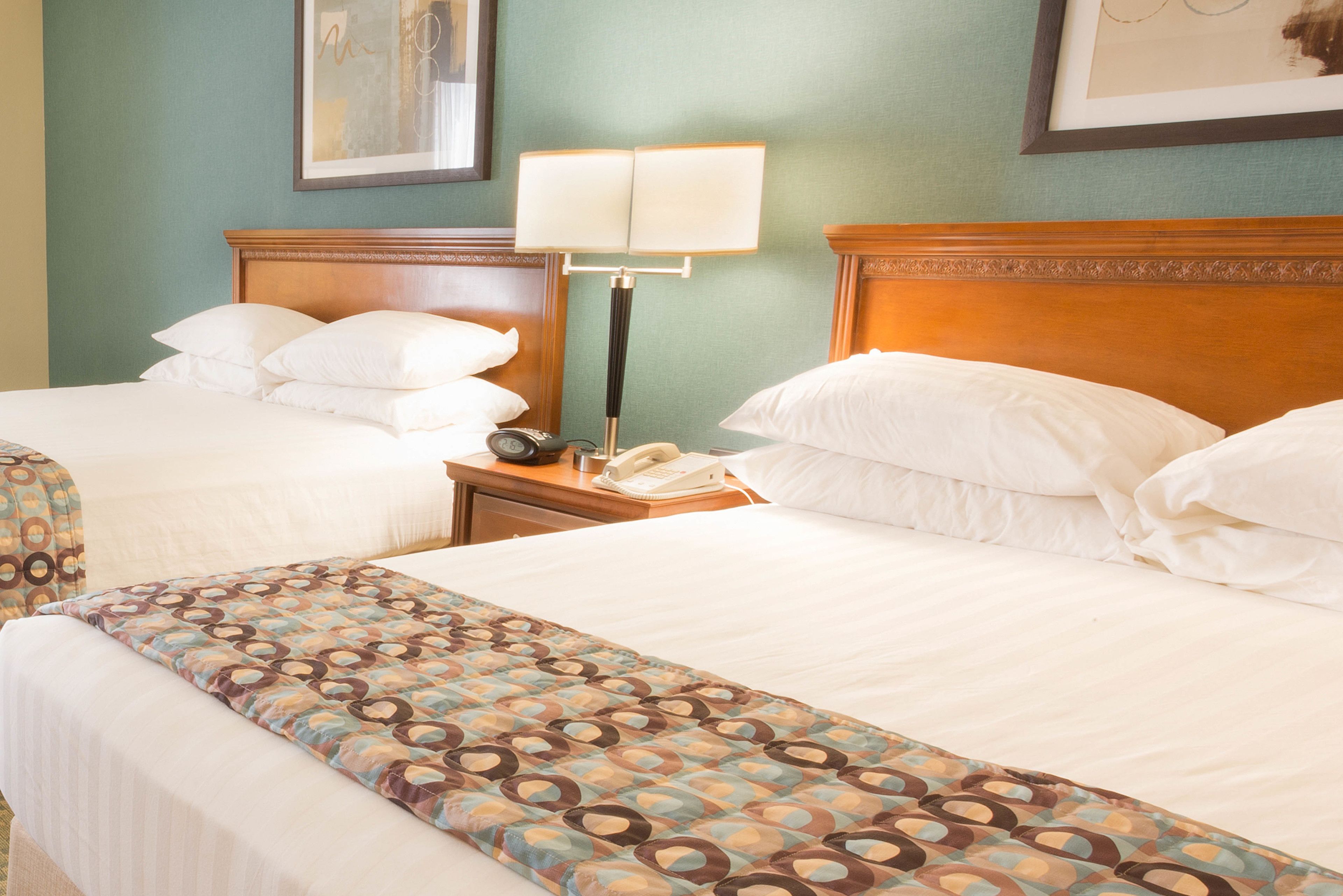 Quarto de hotel com duas camas, uma mesinha de cabeceira com abajur e um quadro na parede.